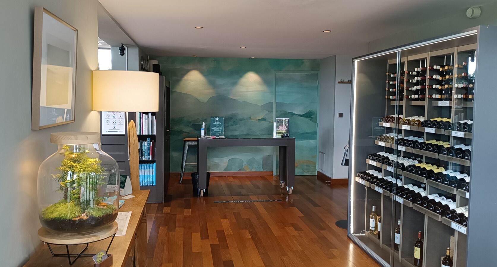 Interior moderno com chão de madeira, uma vitrine de vidro com garrafas de vinho, uma escrivaninha com abajur e uma estante de livros.