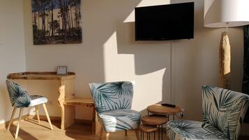 Um quarto aconchegante com uma escrivaninha de madeira, duas cadeiras com padrão de folha tropical, uma mesa redonda de madeira, uma TV fixada na parede e um abajur com base de madeira.