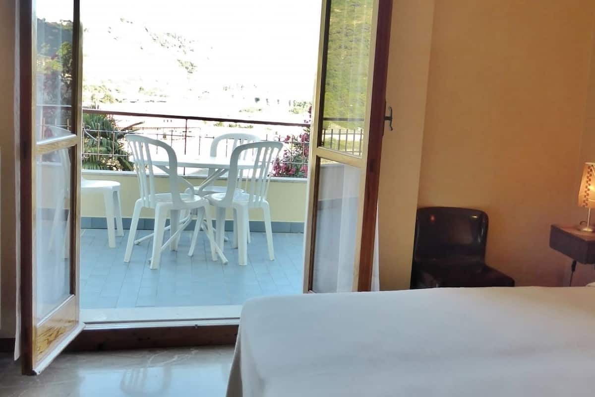 Quarto de hotel com cama, mesa de cabeceira com luminária, cadeira e vista para uma varanda externa com cadeiras e mesa.