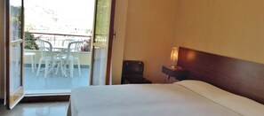 Quarto de hotel com cama, mesa de cabeceira com luminária, cadeira e vista para uma varanda externa com cadeiras e mesa.