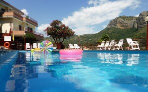 Piscina com água azul cristalina, rodeada por espreguiçadeiras brancas e uma bola de praia colorida. Uma montanha com vegetação é visível ao fundo.