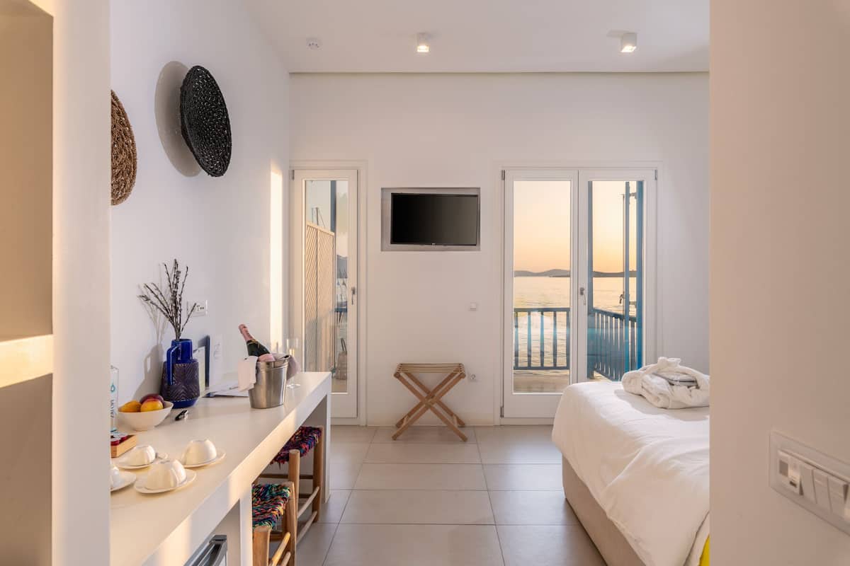 Quarto de hotel com cama, televisão na parede, mesinha com vaso e frutas, e varanda com vista.