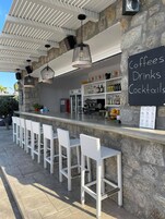 Uma área de bar ao ar livre com banquetas de vime branco, parede de pedra e cardápio em lousa com cafés, bebidas e coquetéis.