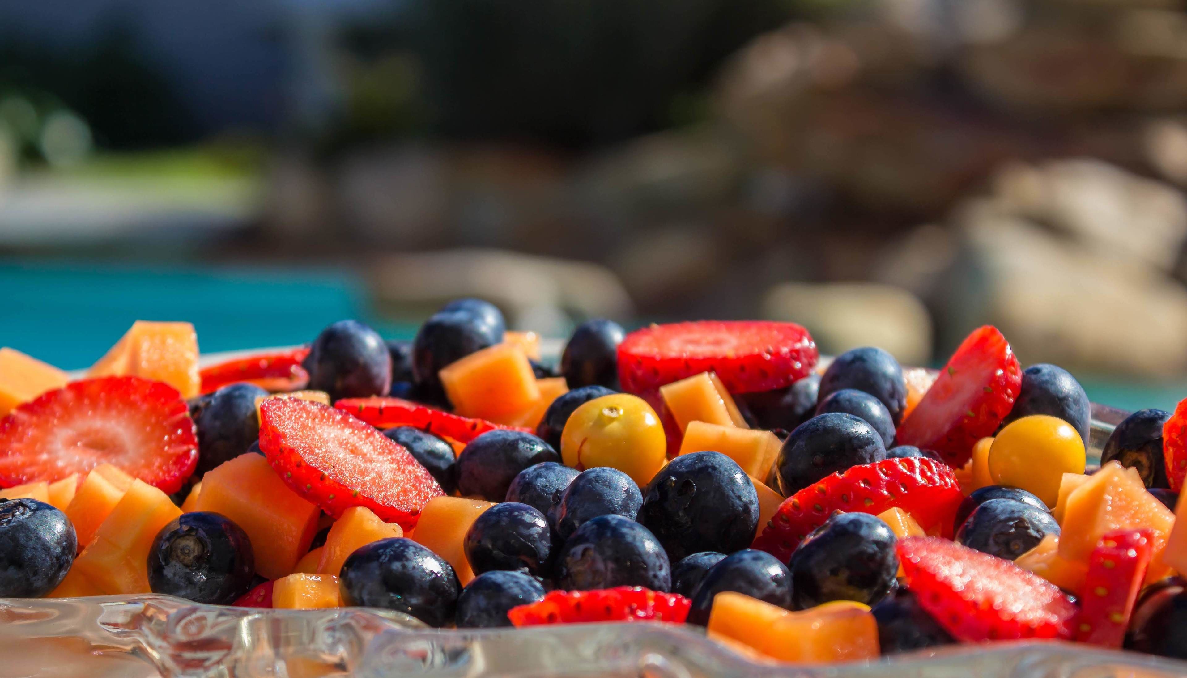 Uma tigela de vidro cheia de frutas frescas variadas, incluindo morangos, mirtilos e melão cantaloupe.