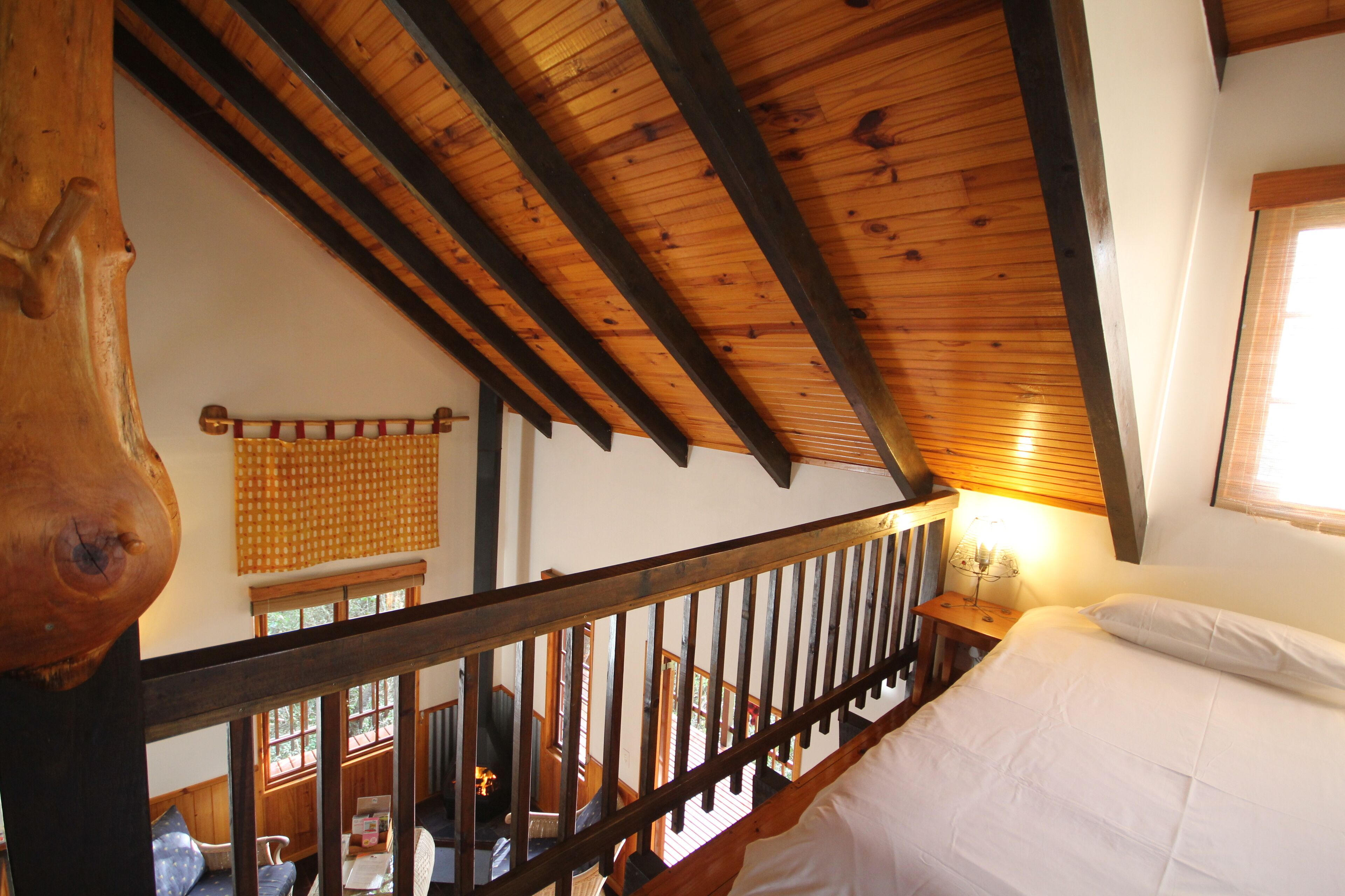 Quarto com teto de madeira, cama com roupa de cama branca e vista para uma área de jantar abaixo.