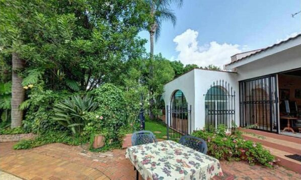 Um pátio com mesa e cadeiras, cercado por vegetação exuberante e um edifício branco com janelas em arco.