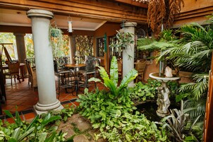 Amplo espaço interno com área de refeições, incluindo mesas e cadeiras, cercado por vegetação exuberante e uma fonte decorativa.