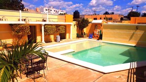 Piscina com área de pátio, rodeada por edifícios com paredes amarelas e um céu azul límpido.