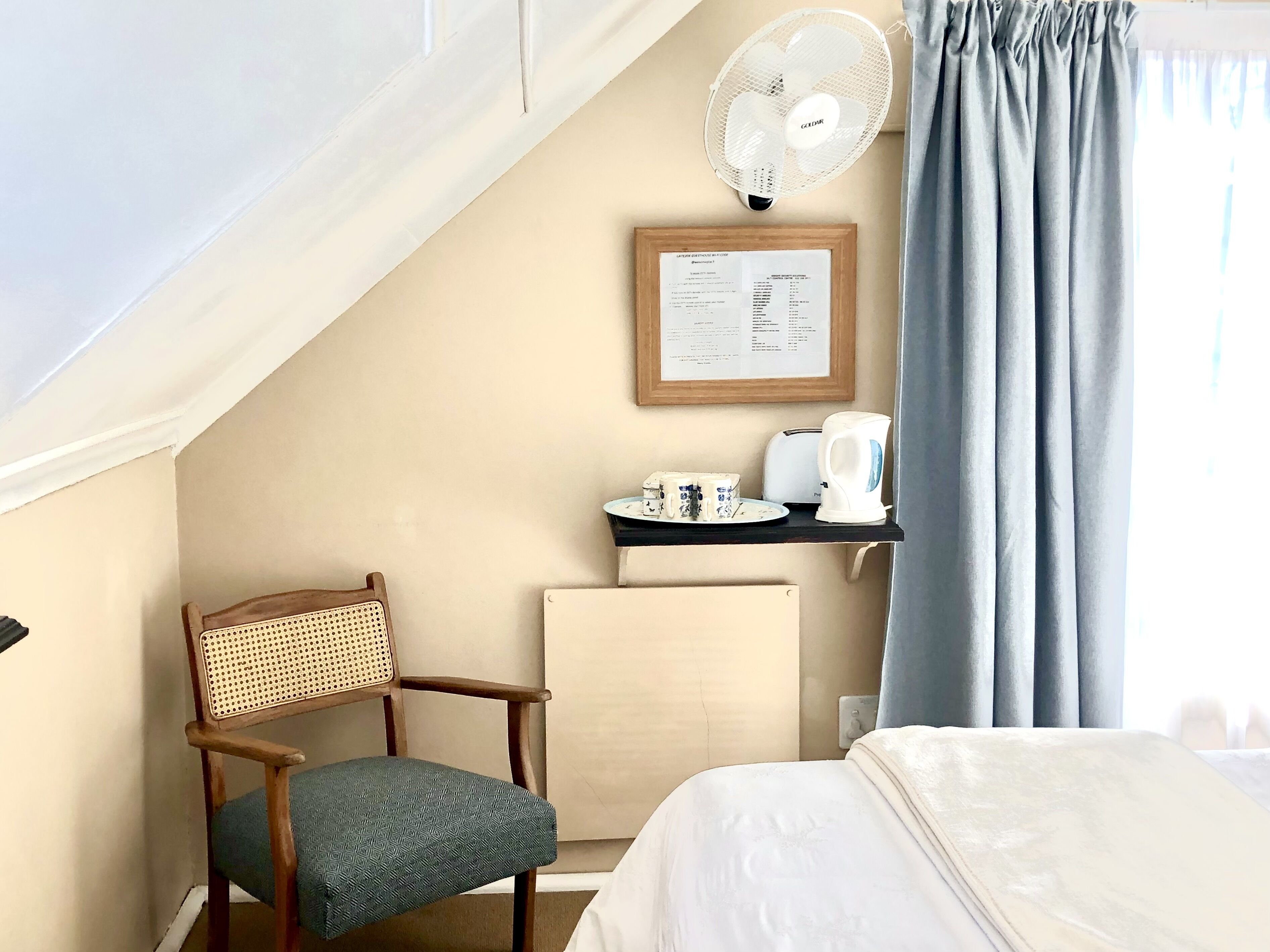 Quarto de hotel com uma cadeira de vime, uma mesa pequena com uma chaleira, um menu emoldurado e uma cama com edredom branco.