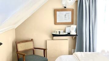 Quarto de hotel com uma cadeira de vime, uma mesa pequena com uma chaleira, um menu emoldurado e uma cama com edredom branco.