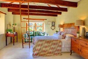 Um quarto aconchegante com teto de madeira, uma escada, uma cama com colcha estampada, mesa de cabeceira com luminária e janela com vista para o verde.