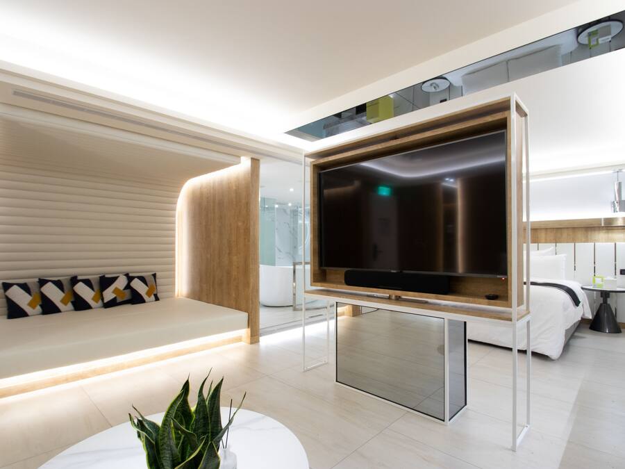 Uma sala de estar moderna com uma grande TV de tela plana fixada em uma estrutura de madeira, um sofá branco com almofadas estampadas, uma mesa de centro de vidro com uma planta e um design minimalista.