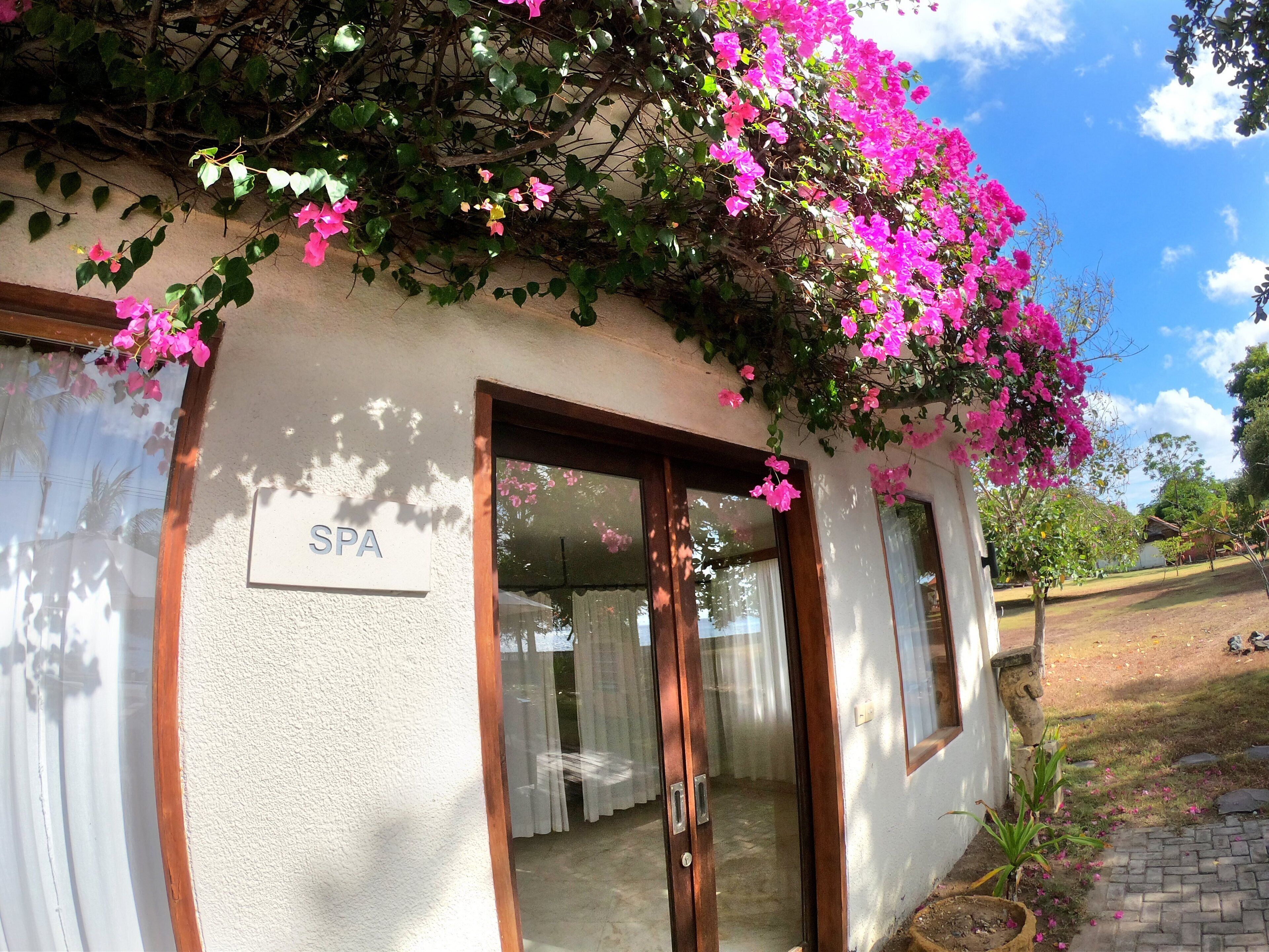 Um edifício de spa com uma placa indicando sua finalidade, rodeado por vegetação exuberante e flores cor-de-rosa.