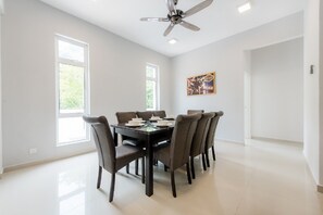 Uma área de refeições com mesa e cadeiras de madeira escura, janelas amplas, ventilador de teto e um quadro na parede.