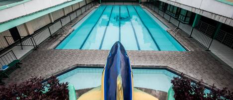 Piscina com prancha de mergulho em primeiro plano, rodeada por uma galeria coberta e um edifício com a placa 'One Way'.