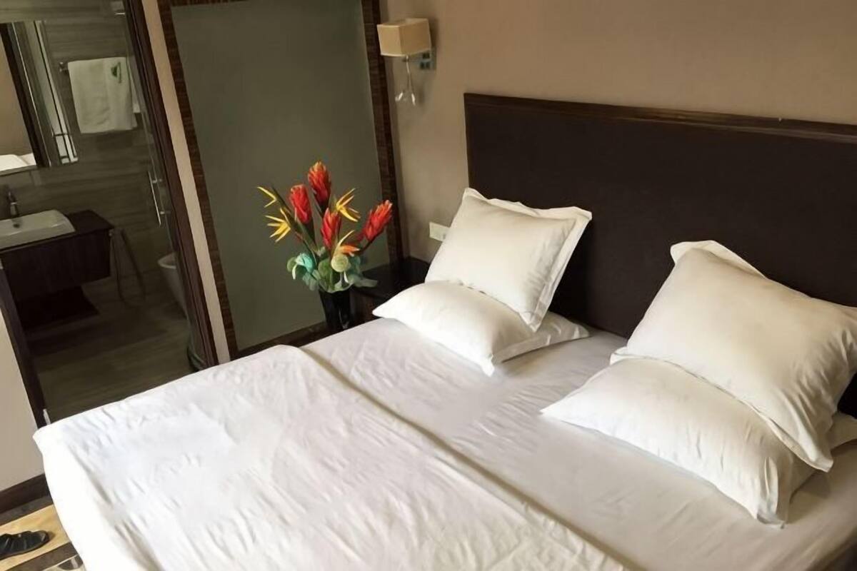 Quarto de hotel com cama, almofadas, mesa de cabeceira, um vaso com flores, um espelho e um banheiro visível através de uma porta de vidro.