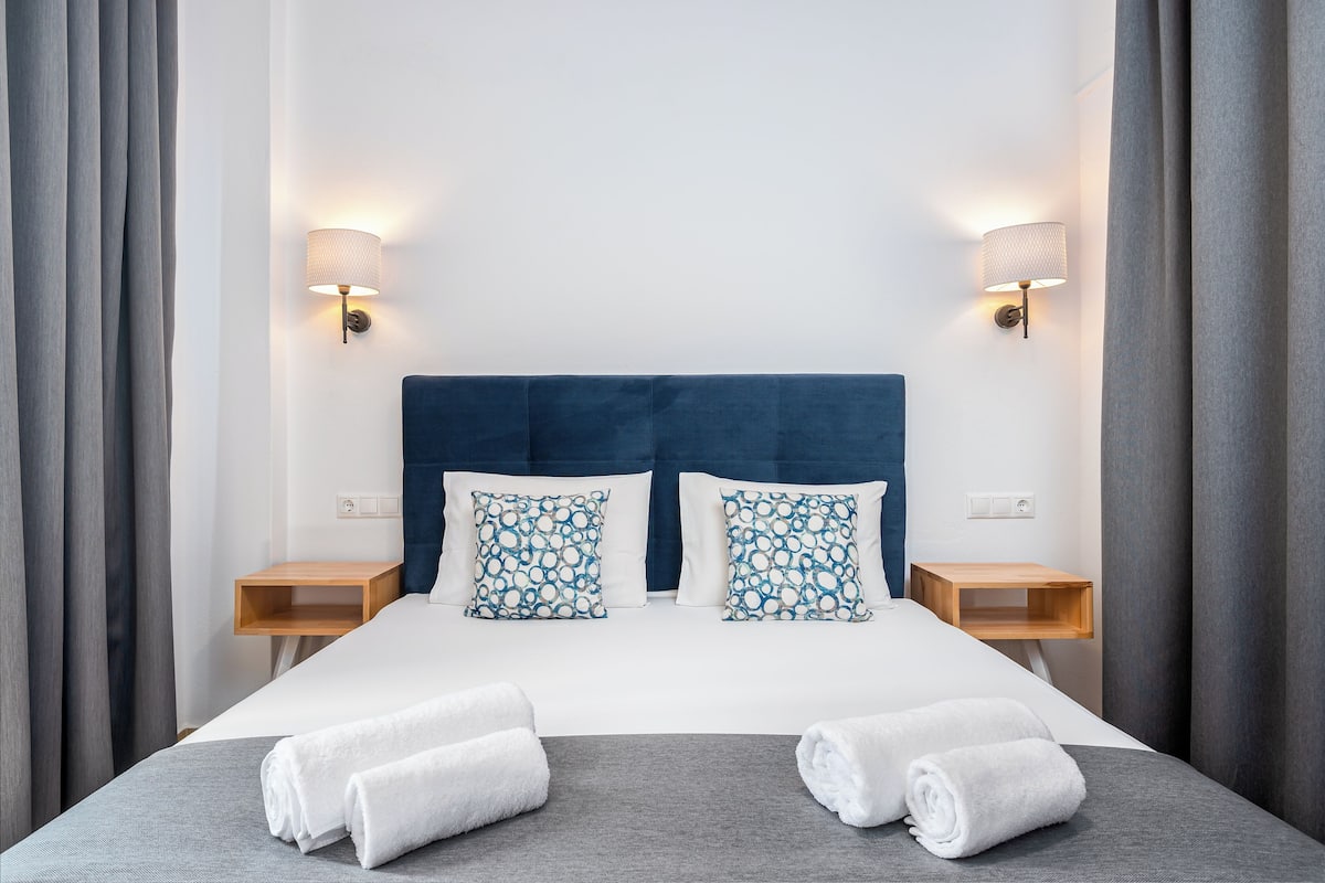 Uma cama bem arrumada com cabeceira azul, duas almofadas iguais, duas mesas de cabeceira com luminárias, e duas toalhas enroladas sobre a cama.