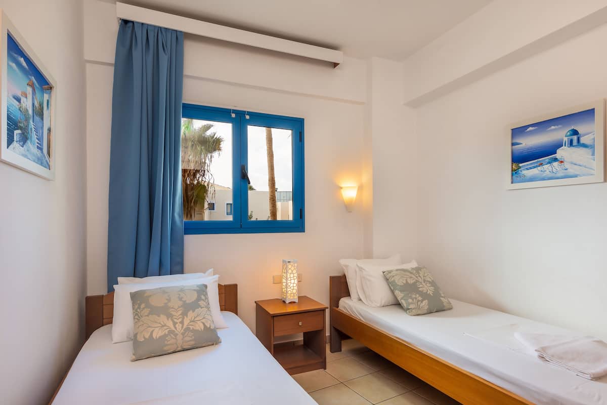 Quarto com duas camas, janela com cortinas azuis, mesa de cabeceira com abajur e luz na parede.