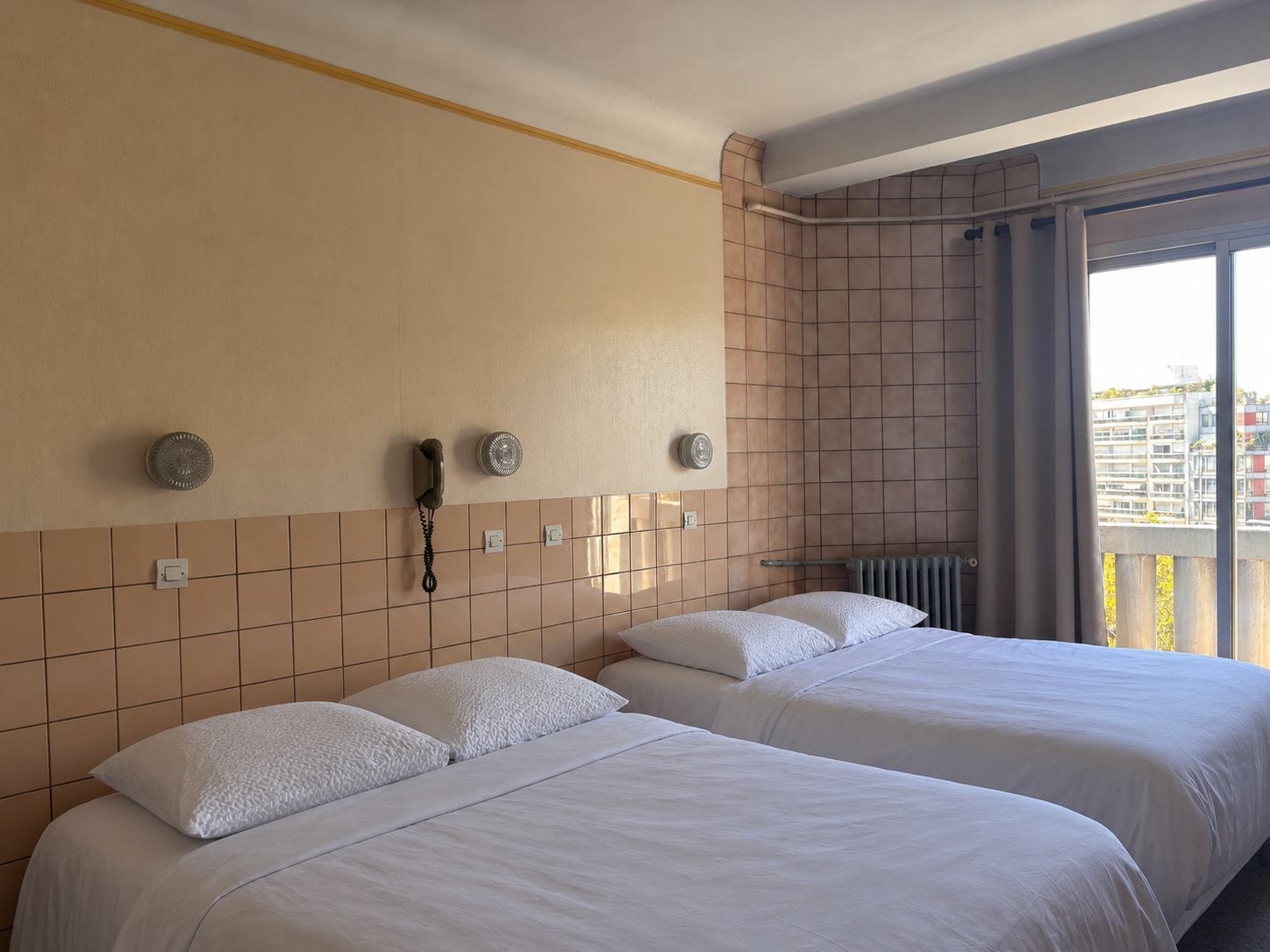 Quarto de hotel com duas camas, telefone e janela com vista para edifícios.