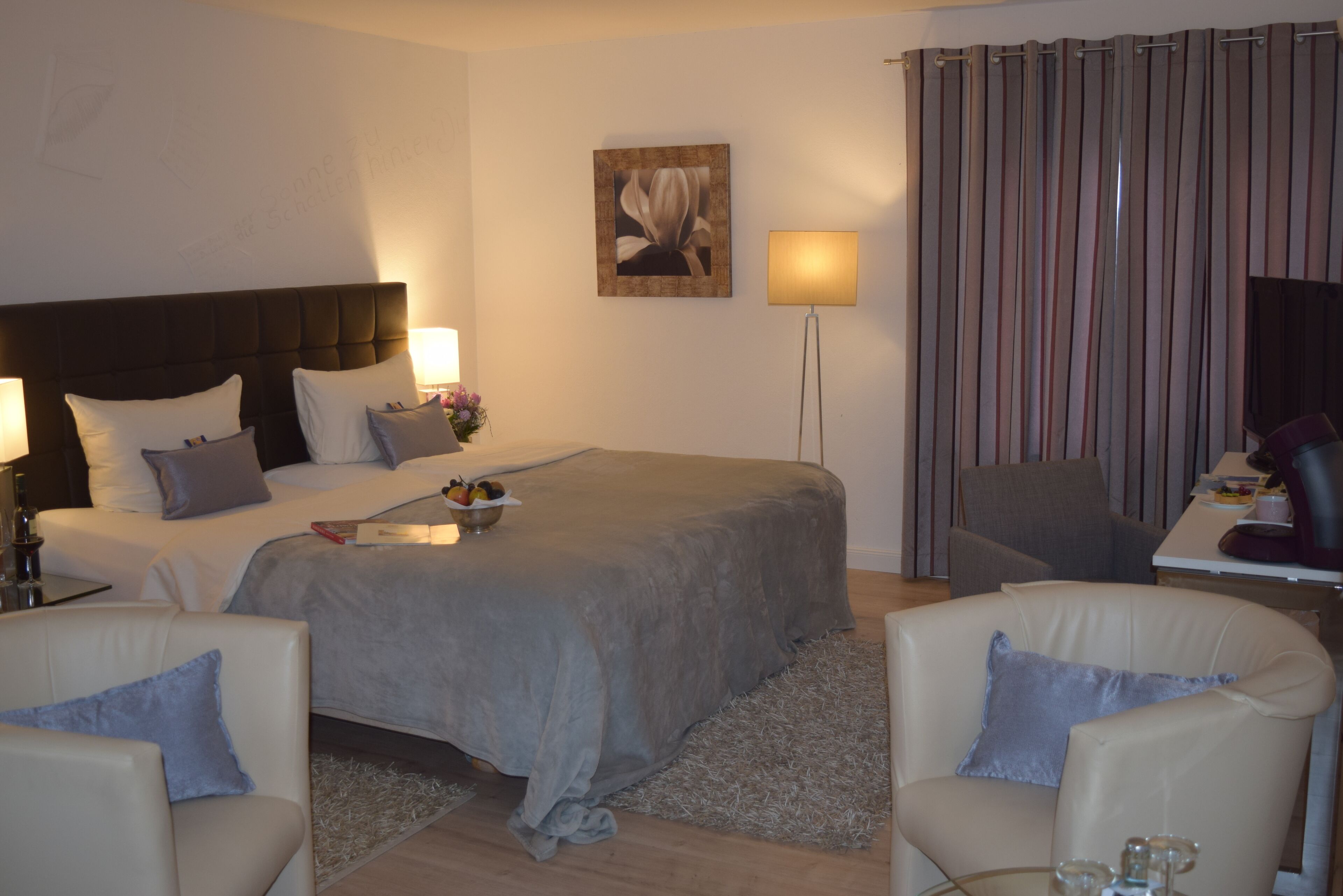 Quarto de hotel com uma cama grande, duas mesas de cabeceira com abajures, uma poltrona, uma escrivaninha com cadeira e uma cortina.