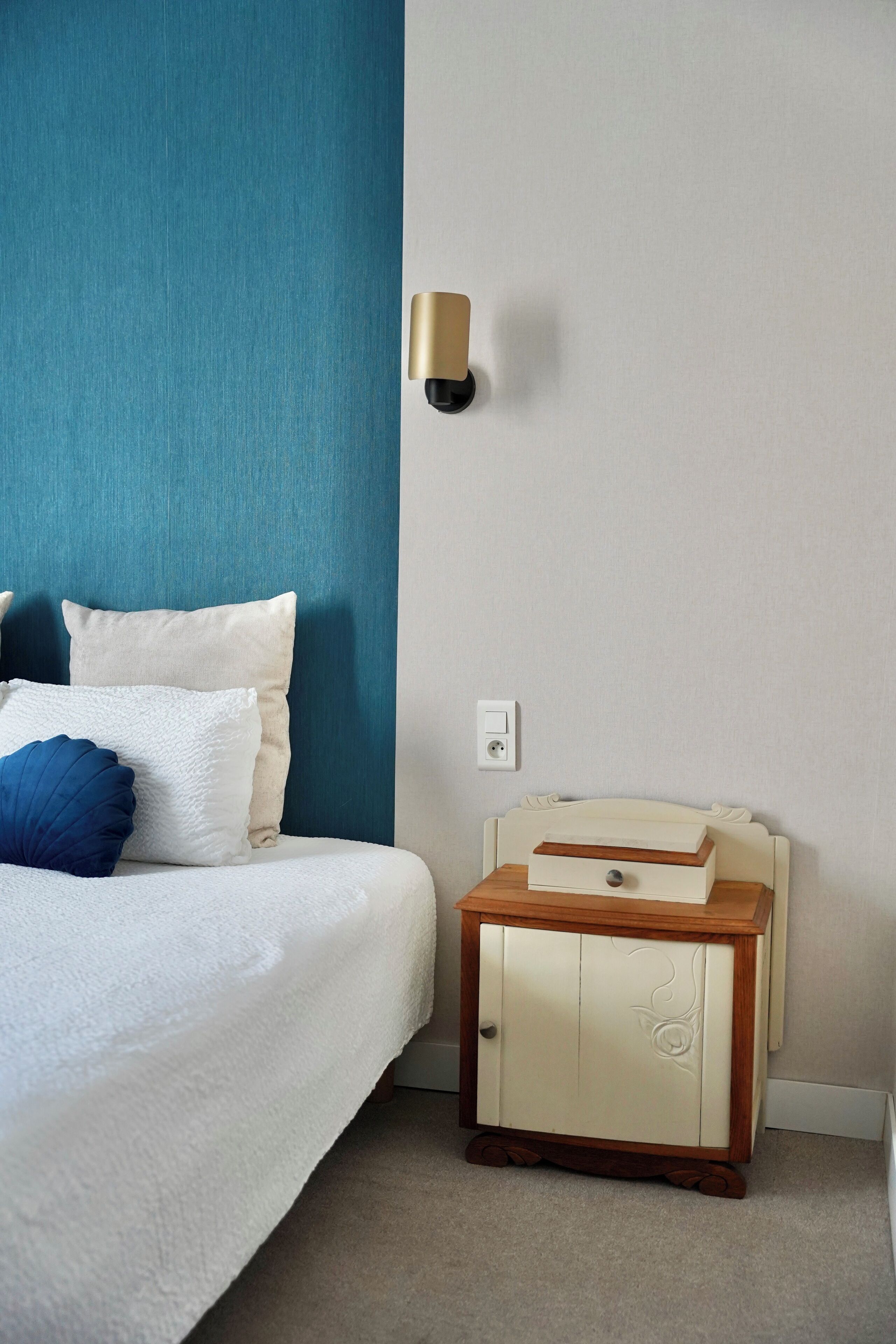 Cama bem arrumada com roupa de cama branca e azul, mesa de cabeceira com gaveta e luminária fixada na parede.