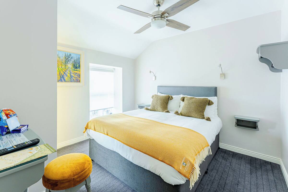 Quarto de hotel com cama, escrivaninha com cadeira, ventilador de teto, janela e um quadro na parede.