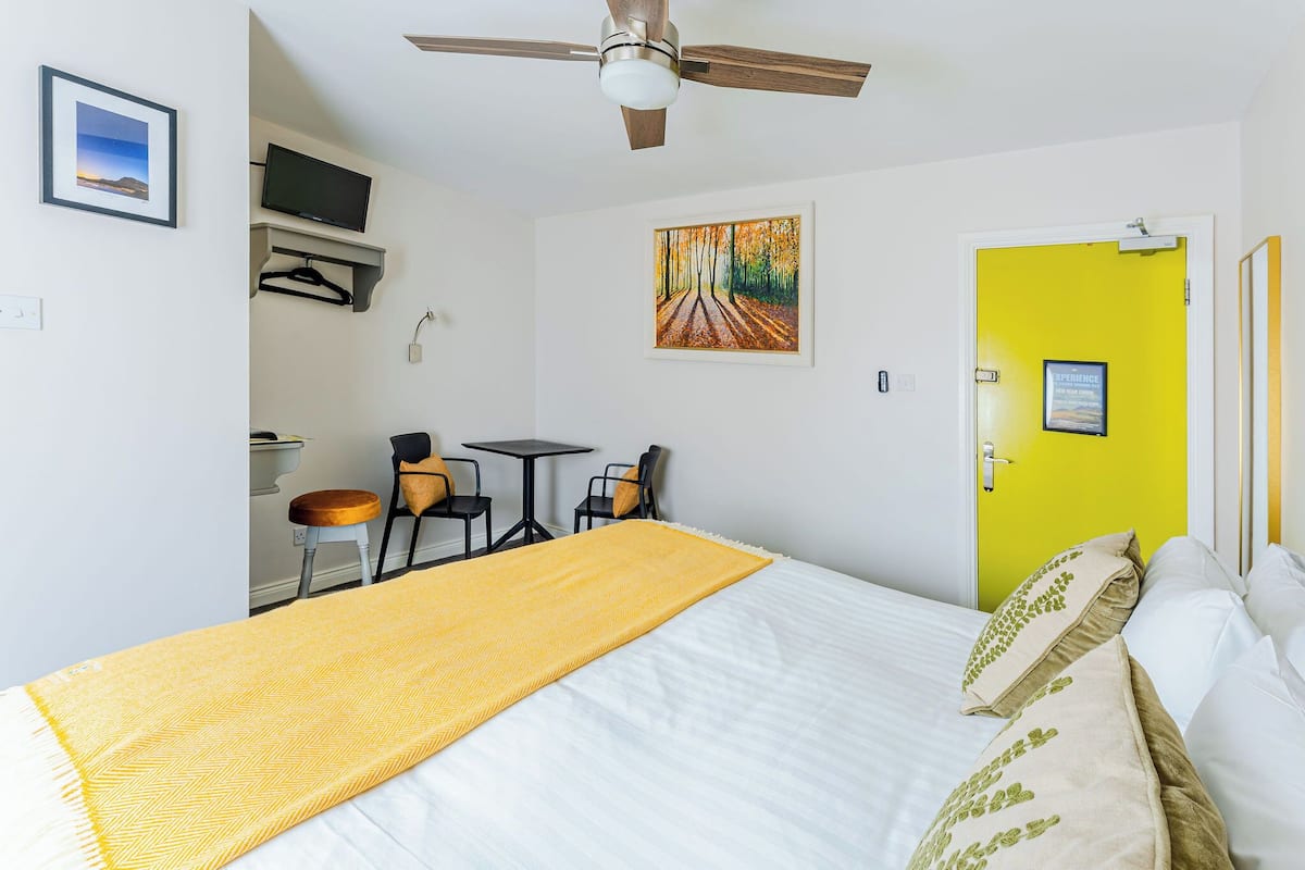 Quarto de hotel com uma cama, um cobertor amarelo, uma escrivaninha com cadeiras, uma televisão e uma porta com uma placa.