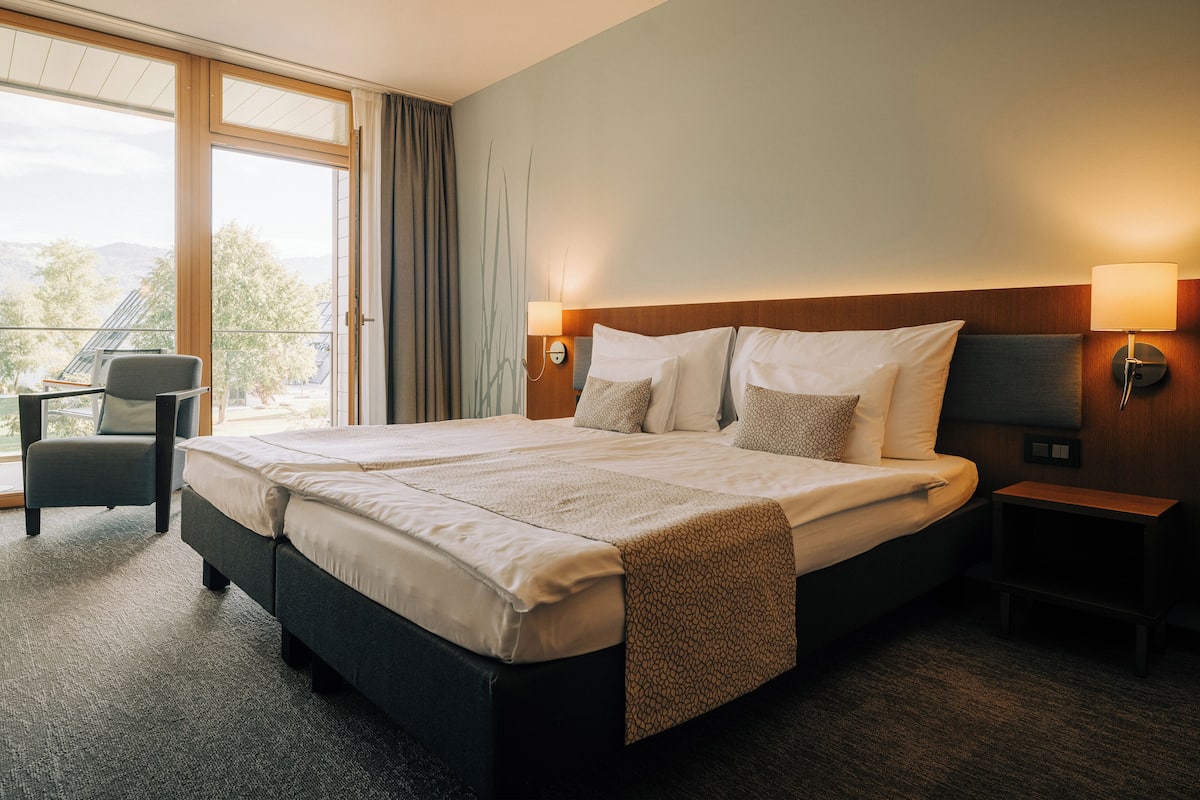 Quarto de hotel com uma cama grande, duas mesas de cabeceira, uma cadeira e vista para o exterior.