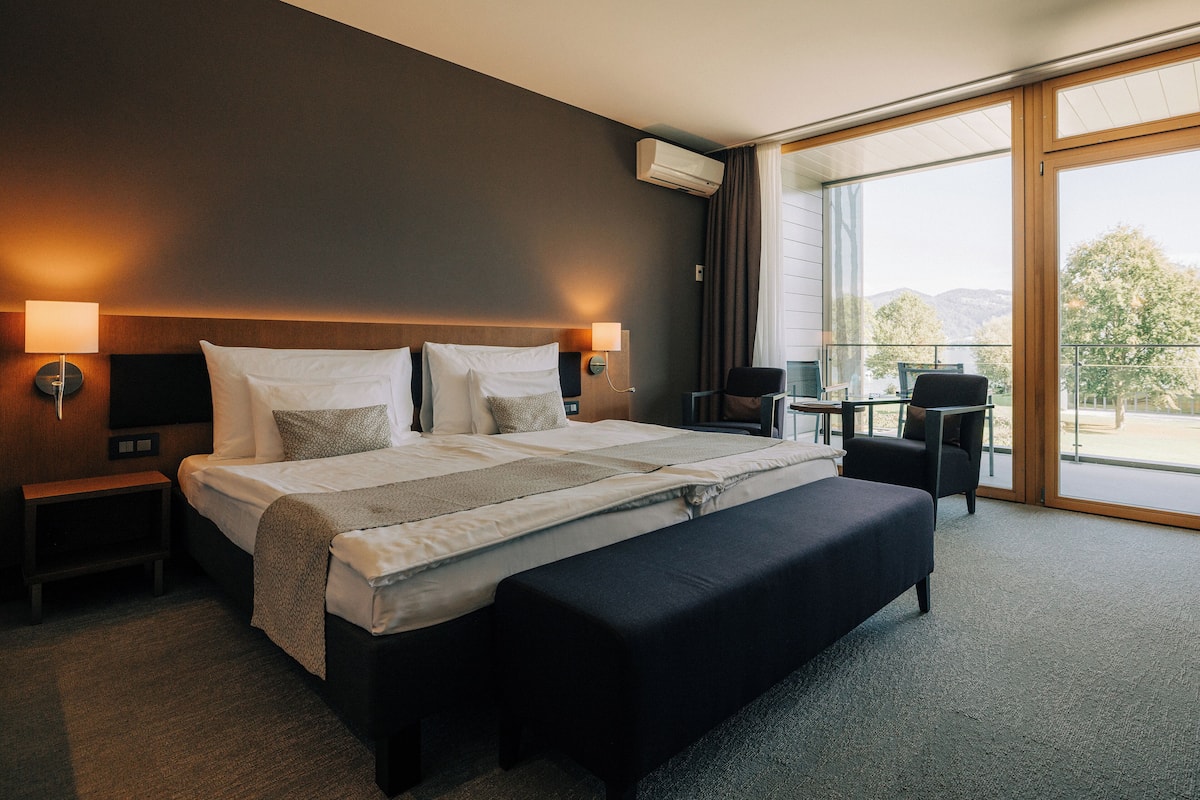 Quarto de hotel com uma cama grande, luminárias laterais, uma mesa de trabalho com uma cadeira e vista para o exterior através de uma grande janela.