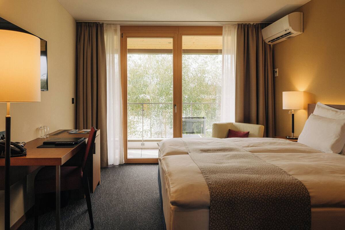 Quarto de hotel com cama, escrivaninha, cadeira, varanda com cortinas e ar condicionado.