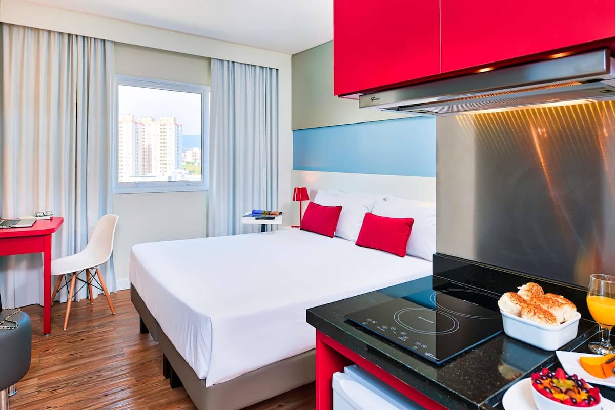 Quarto de hotel com cama, escrivaninha, cadeira, janela com cortinas, cozinha compacta com fogão e disposição para café da manhã.