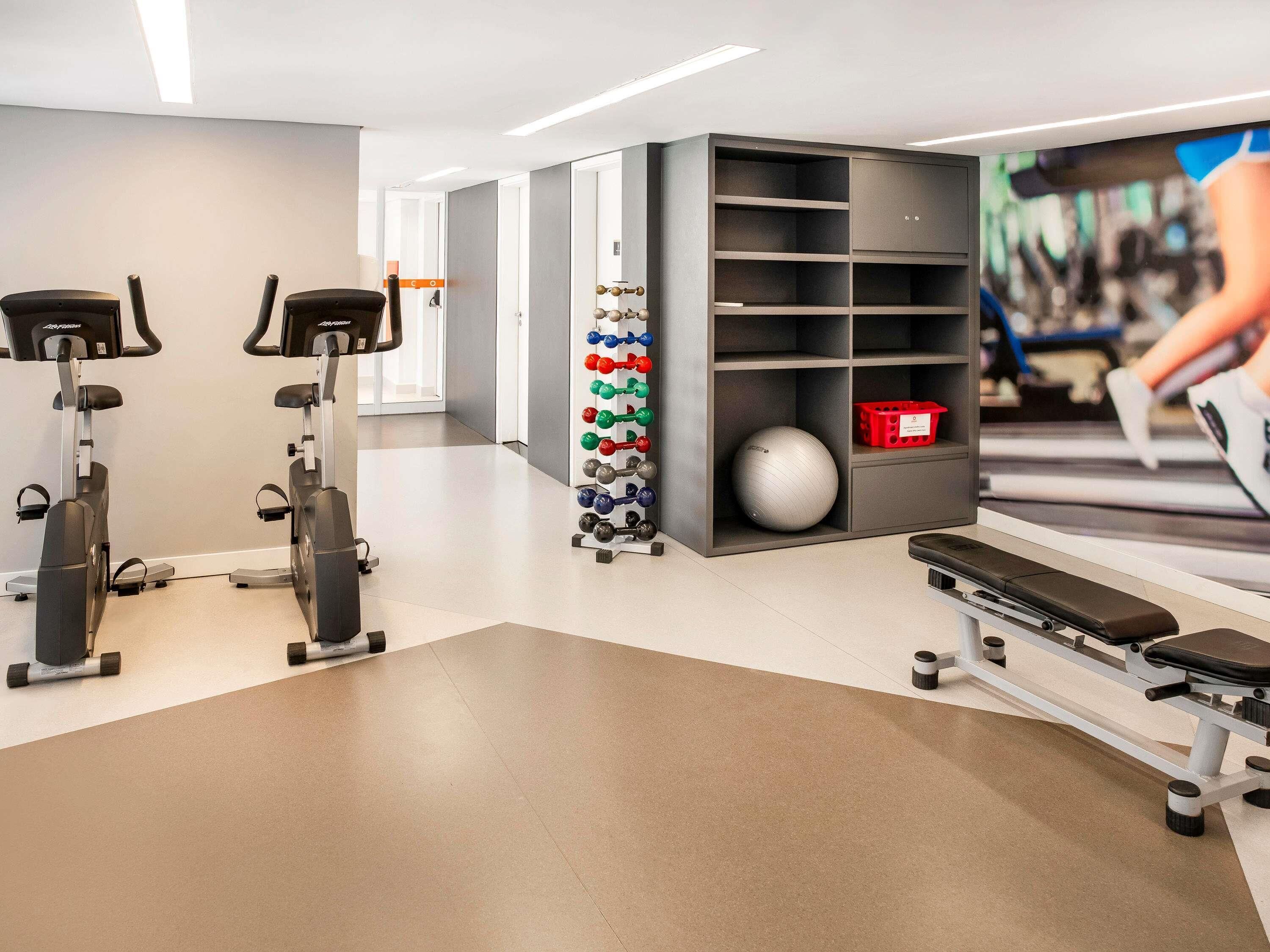 Uma academia com bicicletas ergométricas, um suporte de halteres na parede, um banco de musculação e uma esteira.