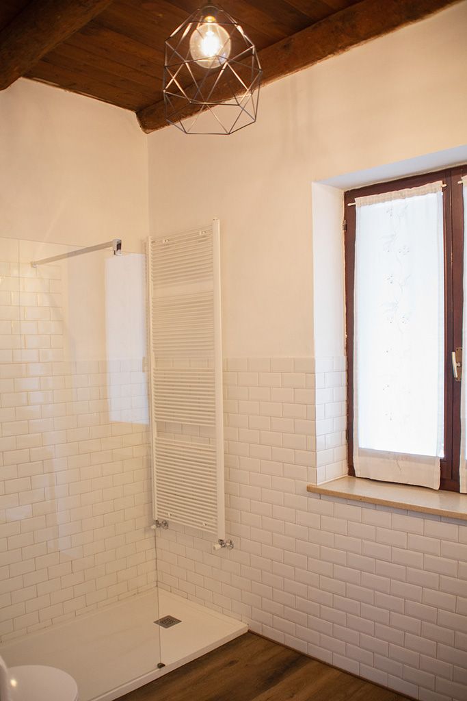 Uma casa de banho com paredes em azulejos brancos, teto de madeira, chuveiro com porta de vidro, um porta-toalhas e uma janela com moldura de madeira.
