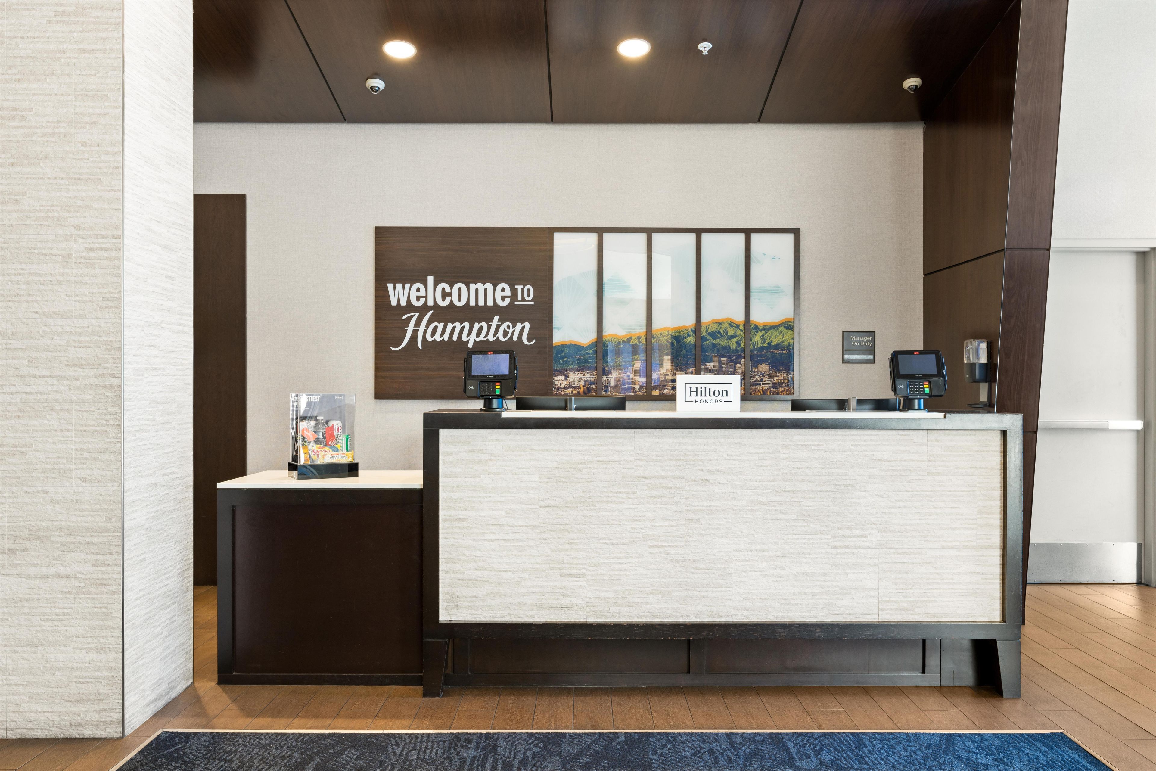 Bancada de recepção com uma placa de 'Welcome to Hampton', vista da cidade e uma placa 'Hilton Honors'.
