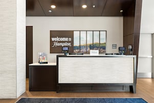 Bancada de recepção com uma placa de 'Welcome to Hampton', vista da cidade e uma placa 'Hilton Honors'.