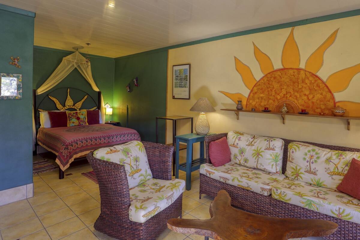 Quarto com cama, sofá, duas cadeiras, mesa de centro de madeira, painel decorativo na parede, prateleira com objetos decorativos e abajur.