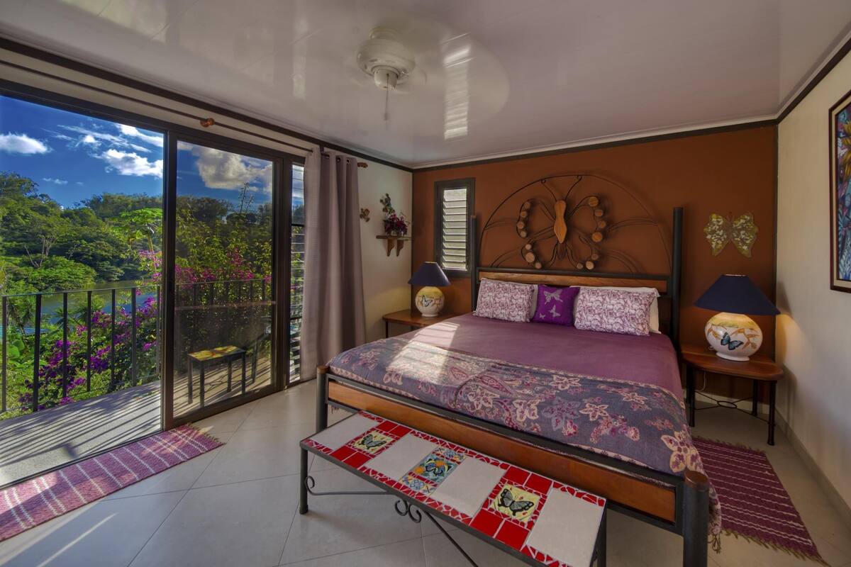 Quarto com cama grande, vista para vegetação exuberante através de porta de vidro deslizante e ventoinha no teto.
