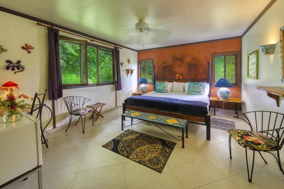 Quarto com cama, duas cadeiras, escrivaninha, ventilador de teto e janelas amplas.
