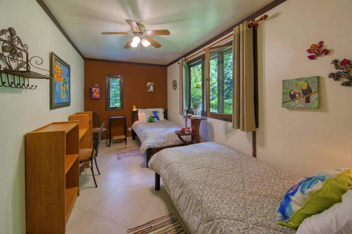 Quarto com duas camas de solteiro, uma escrivaninha, uma cadeira e um ventilador de teto.