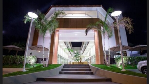 Um edifício moderno com entrada central, ladeado por palmeiras e iluminado por postes de luz.