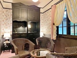 Um quarto com um armário de madeira escura, cadeiras de vime, uma mesa de vidro e uma janela com cortinas.
