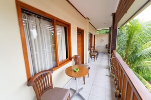 Varanda com guarda-corpo de madeira, mesa e cadeiras. Há janelas com cortinas, uma porta e vista para o verde lá fora.