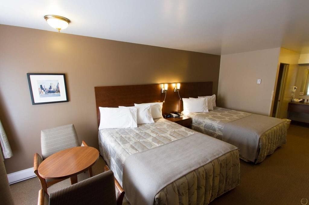 Quarto de hotel com duas camas, uma mesinha e uma cadeira. Há um quadro na parede e uma mesa de cabeceira com abajur.