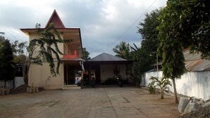 Um edifício de dois andares com telhado vermelho e varanda. Há sacos de cimento no chão e uma árvore em primeiro plano.