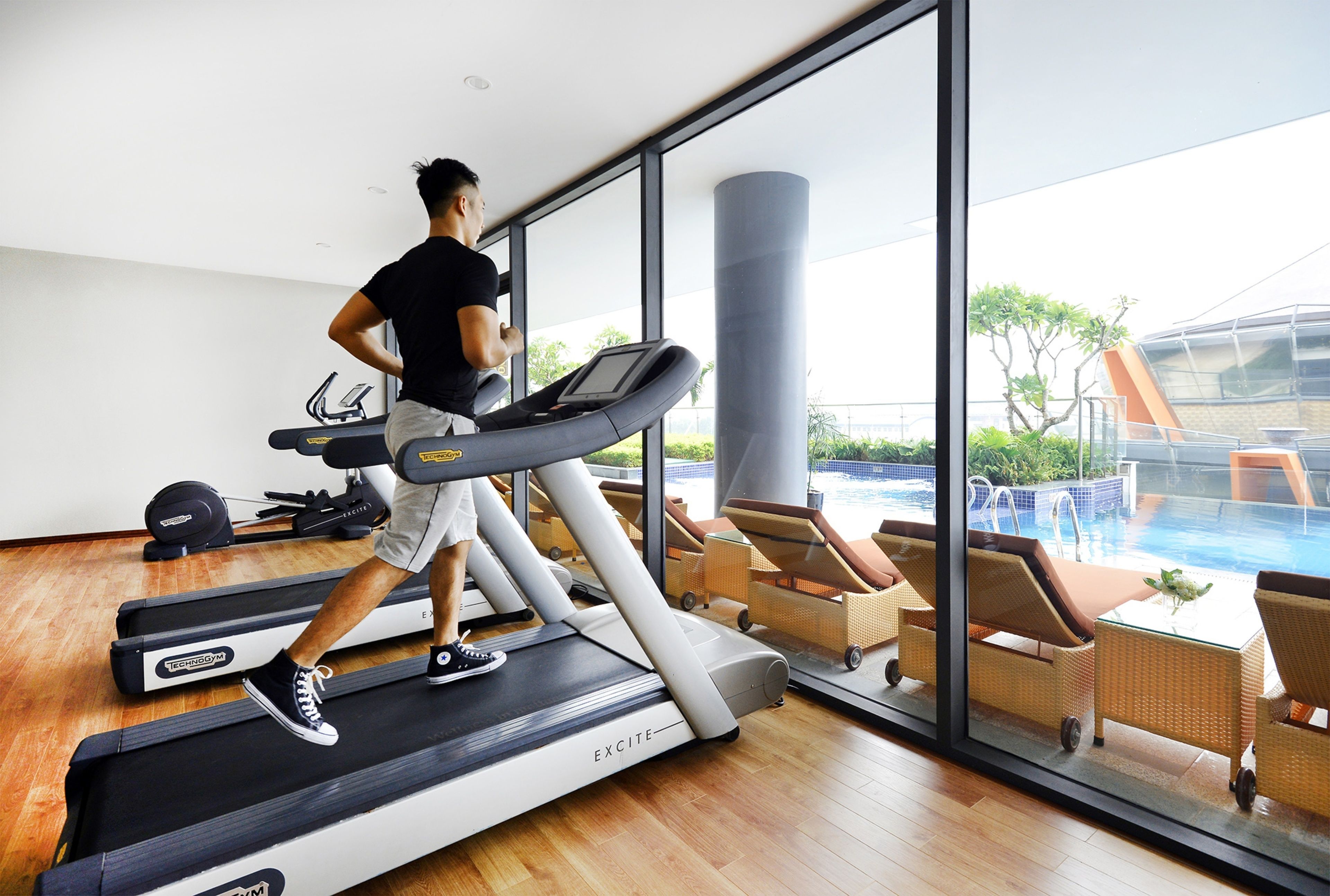 Uma pessoa correndo numa esteira dentro de um quarto de hotel moderno, com vista para uma piscina e área externa com mobiliário.