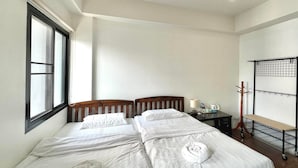 Quarto de hotel com duas camas, uma janela, um cabideiro e uma pequena mesa com abajur.