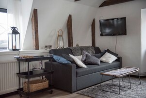 Uma sala de estar moderna com um sofá cinzento, uma TV fixada na parede e teto com vigas de madeira.
