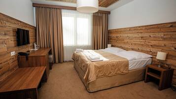 Quarto de hotel com cabeceira de madeira, cama com colcha bege, mesa de cabeceira com abajur, escrivaninha com televisão de tela plana e uma grande janela com cortinas.