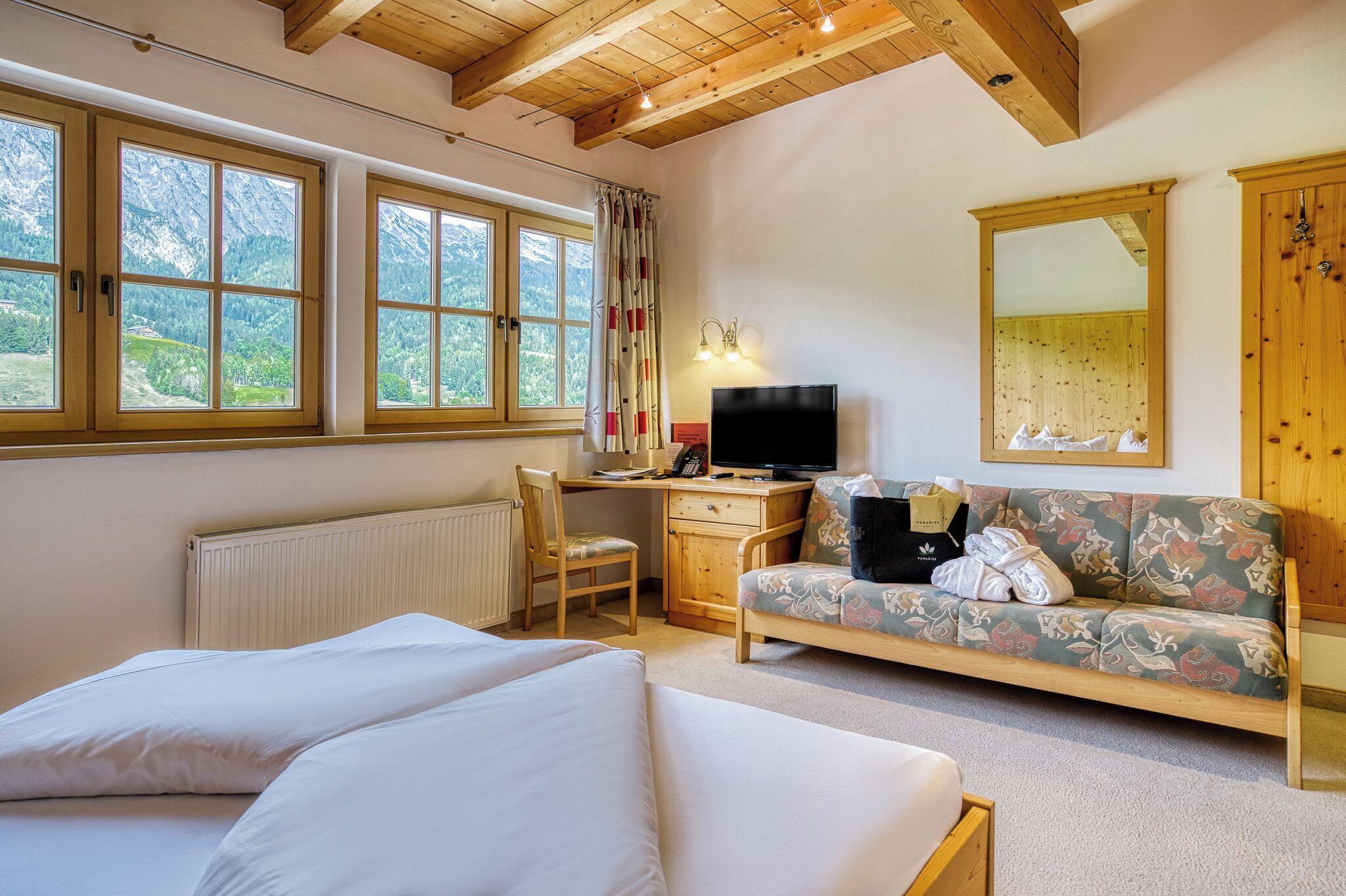 Quarto com duas camas, uma escrivaninha, uma cadeira, um sofá, uma televisão e janela com vista para as montanhas.