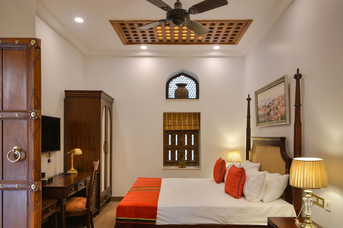 Quarto de hotel com cama, escrivaninha, cadeira, ventilador de teto e janela com design decorativo.